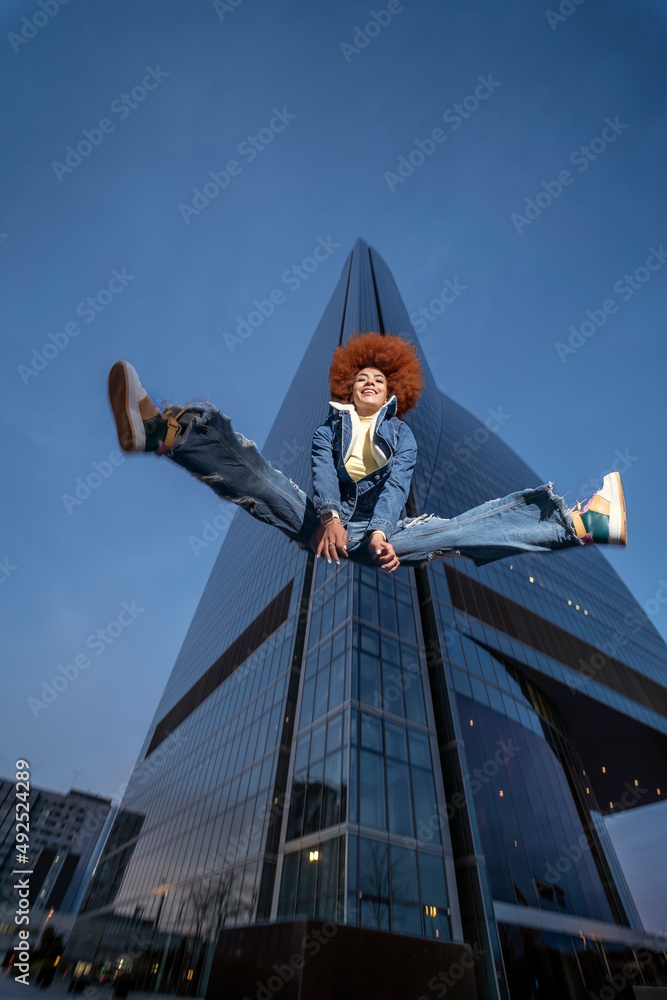 Smiling woman in redhead Afro hairstyle jumping in front of Skyscraper ...