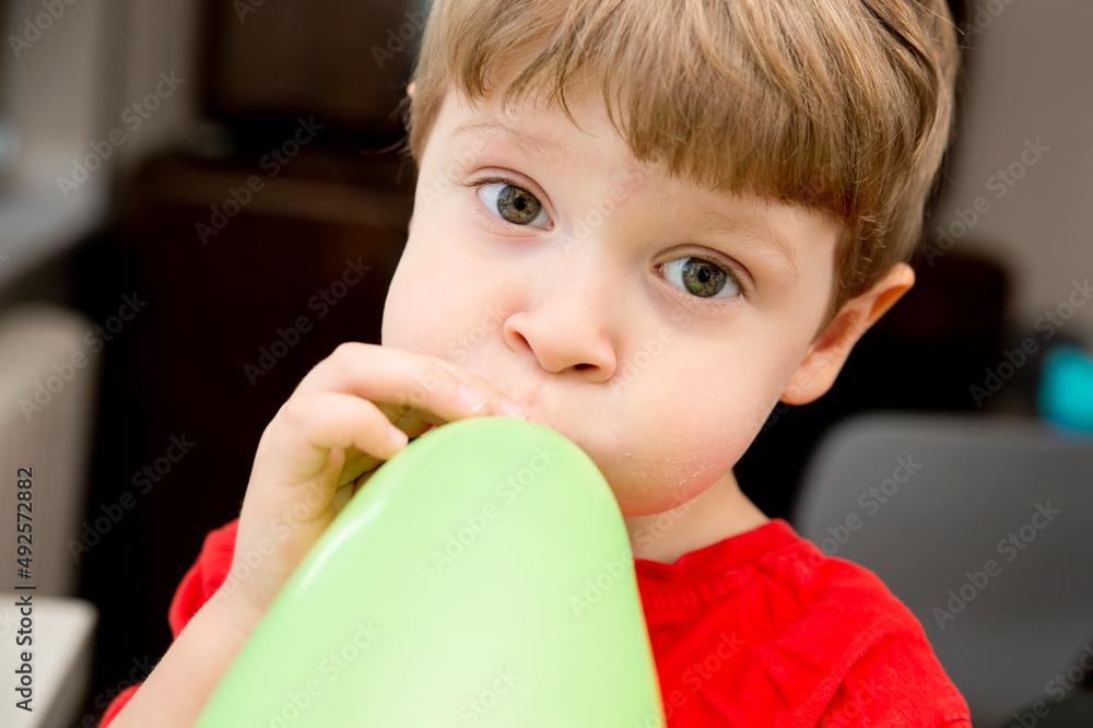 3 year old boy inflating green balloon with a mouth. Swollen cheeks and ...