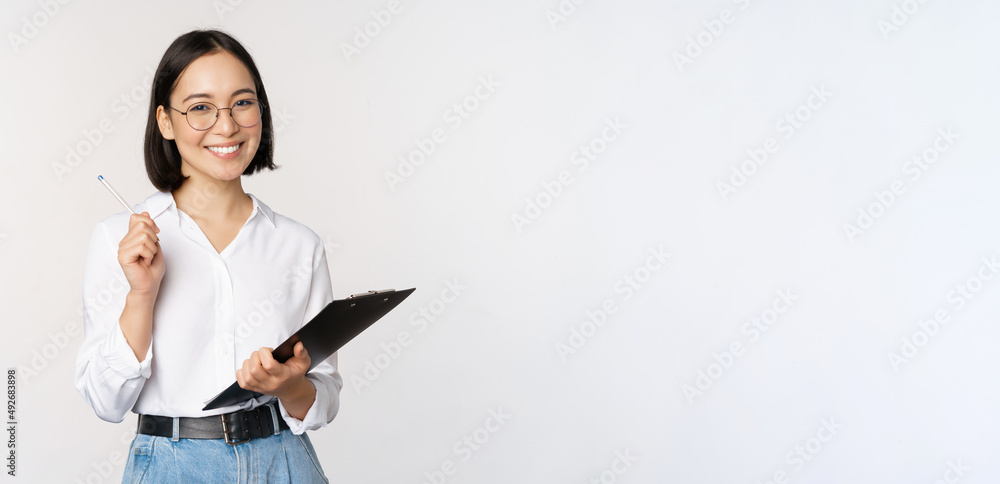 Smiling young asian woman taking notes with pen on clipboard, looking ...
