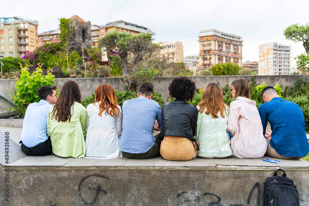 Multi ethnic young people sitting on a bench in a park from behind ...