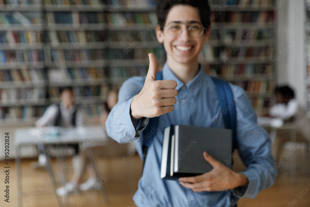Smiling student guy in glasses holds books and textbooks showing thumbs up gesture at camera ...