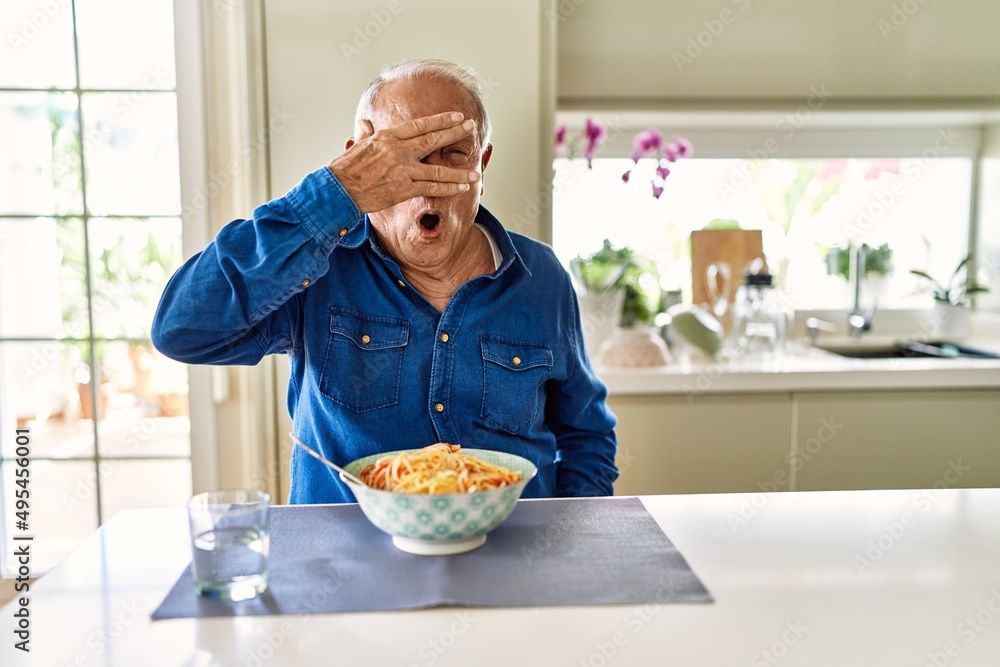 Senior man with grey hair eating pasta spaghetti at home peeking in ...