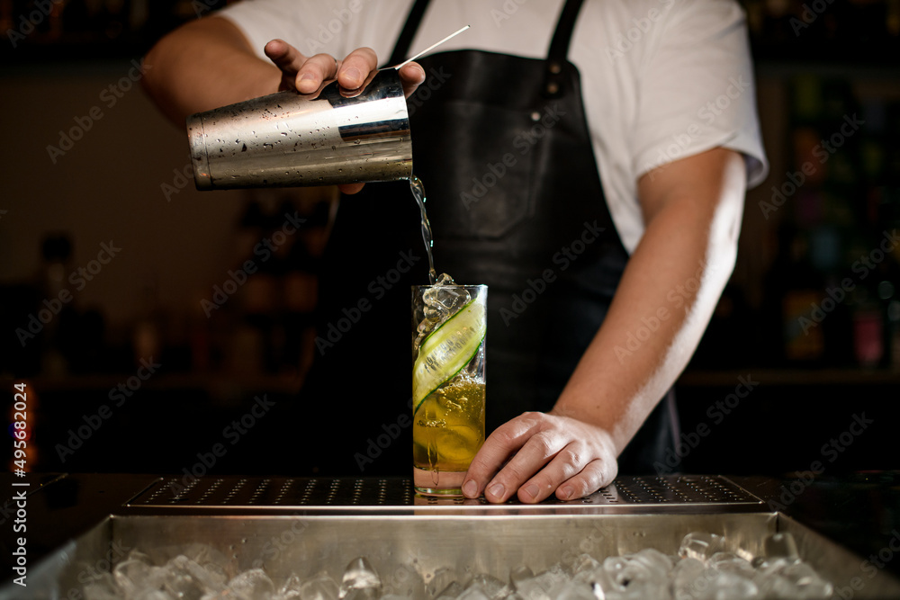 male hand holds shaker and pours drink into glass with cucumber slices ...