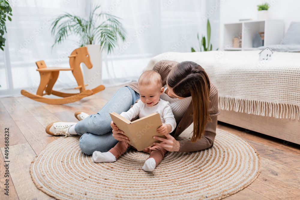 Brunette mom reading book near cheerful baby on rug at home. Stock ...