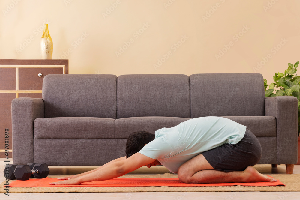 Portrait of a young man doing child pose yoga on exercise mat at home ...