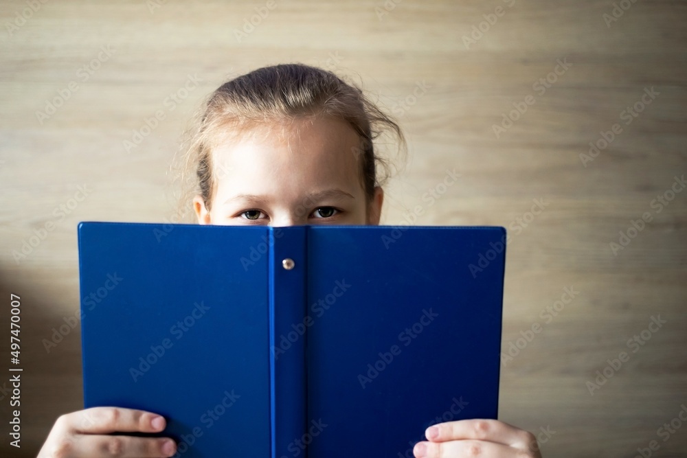 little girl child reads a blue book and looks out from behind it, eyes ...