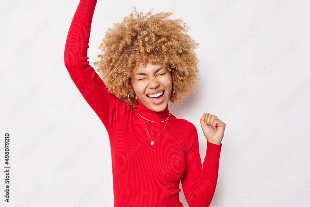 Cheerful happy young woman with curly hair dances and raises arms feels ...