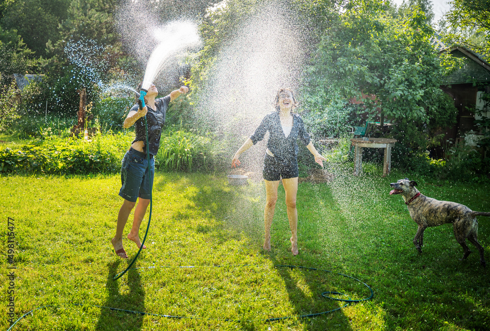 Two beautiful young cheerful woman pouring water from a sprinkler to herself in a green garden ...