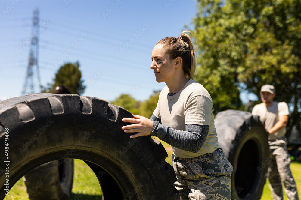 Caucasian female soldier rolling a tire during obstacle course at boot ...
