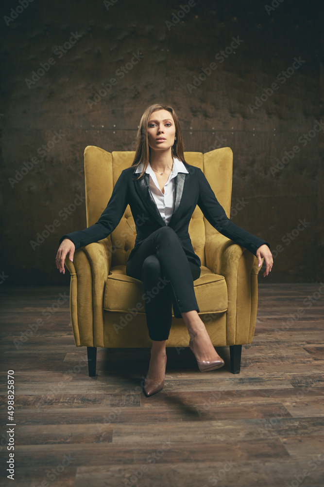 Beautiful young stylish woman in black suit sitting in yellow armchair ...
