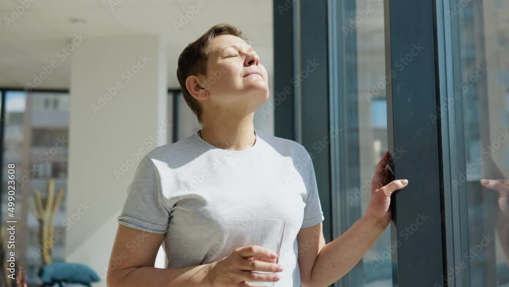 Senior woman drinks water standing near the window in a light room ...