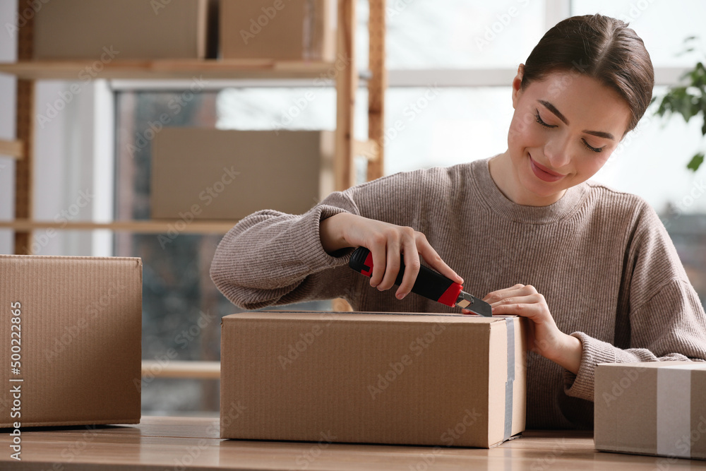 Young woman using utility knife to open parcel at wooden table indoors ...