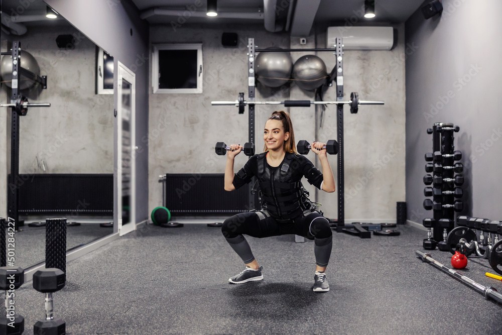 A young and healthy woman in a special black EMS suit performs squats ...