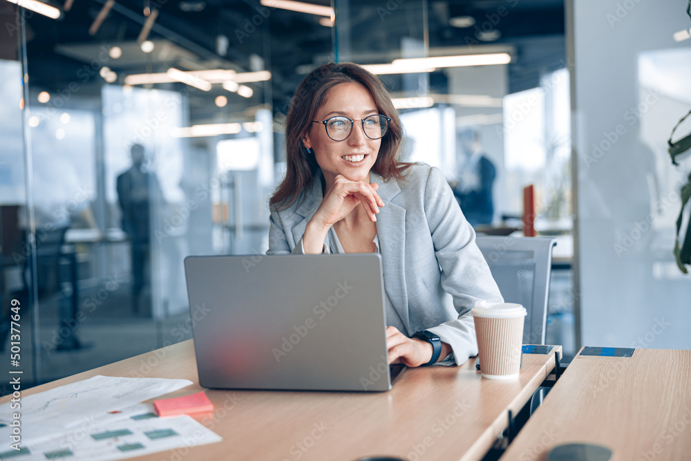 Smiling businesswoman with glasses working on laptop and looking out ...
