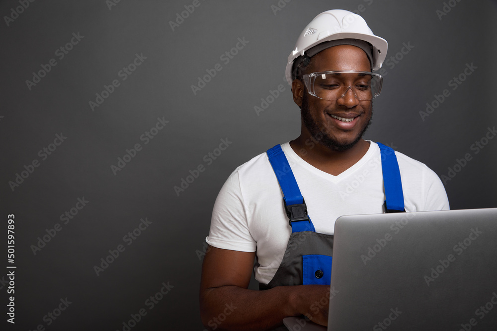 A handsome black man in a white helmet and overalls with a smile works ...