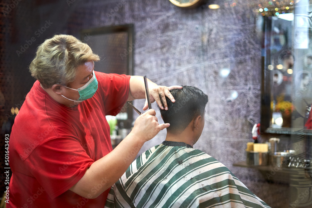 Fat concentrated barber cutting the hair of an asian client in a barber ...
