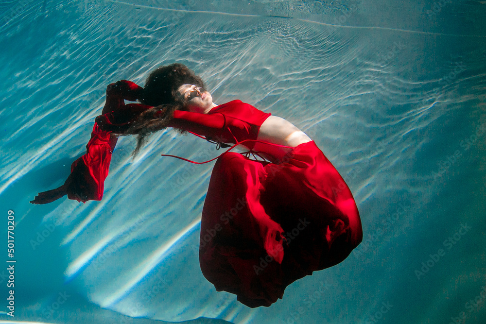 Attractive red-haired young woman swims beautifully underwater in a red ...