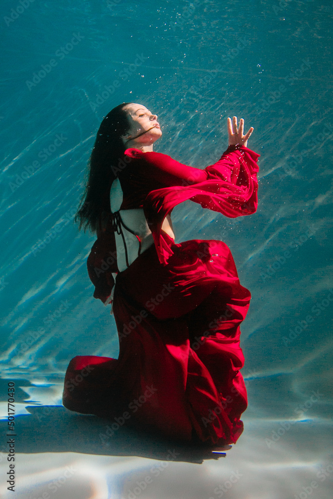 Attractive red-haired young woman swims beautifully underwater in a red ...