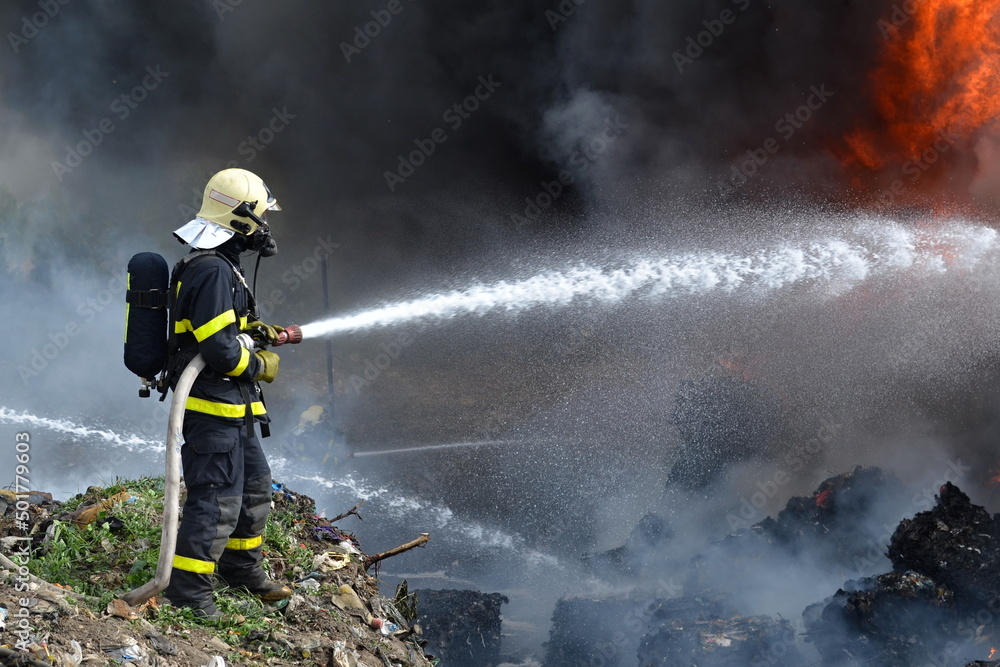 A fireman extinguishes huge a landfill fire with flames and black smoke ...