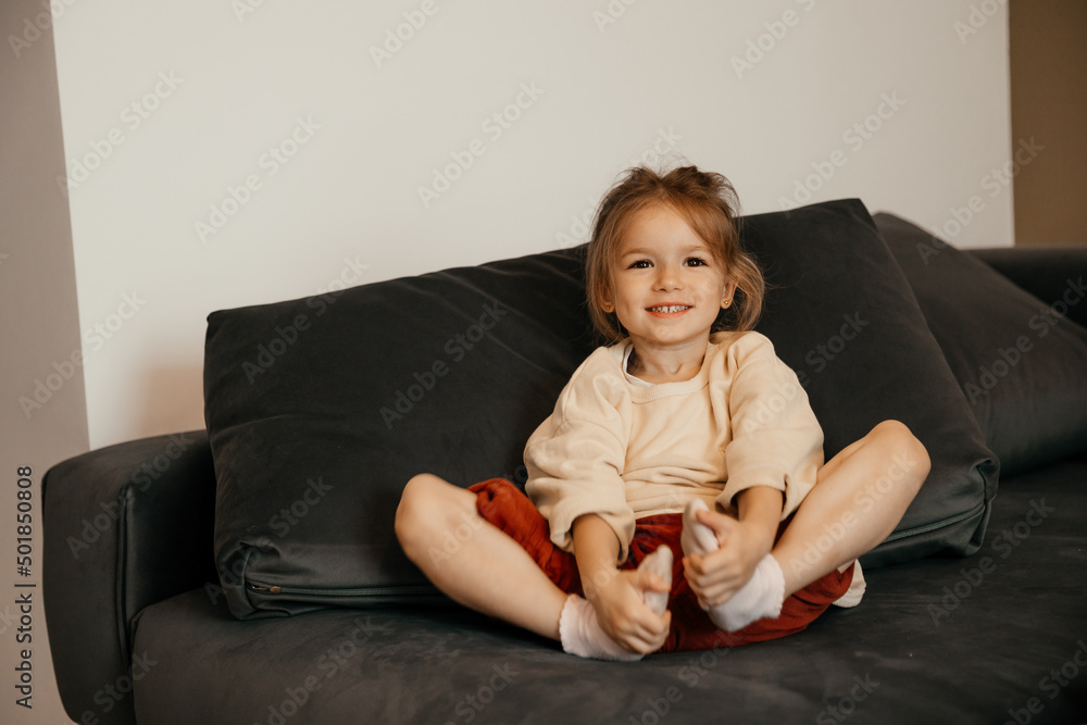 little smiling girl sitting on the couch holding herself by the feet with a smile at the camera ...