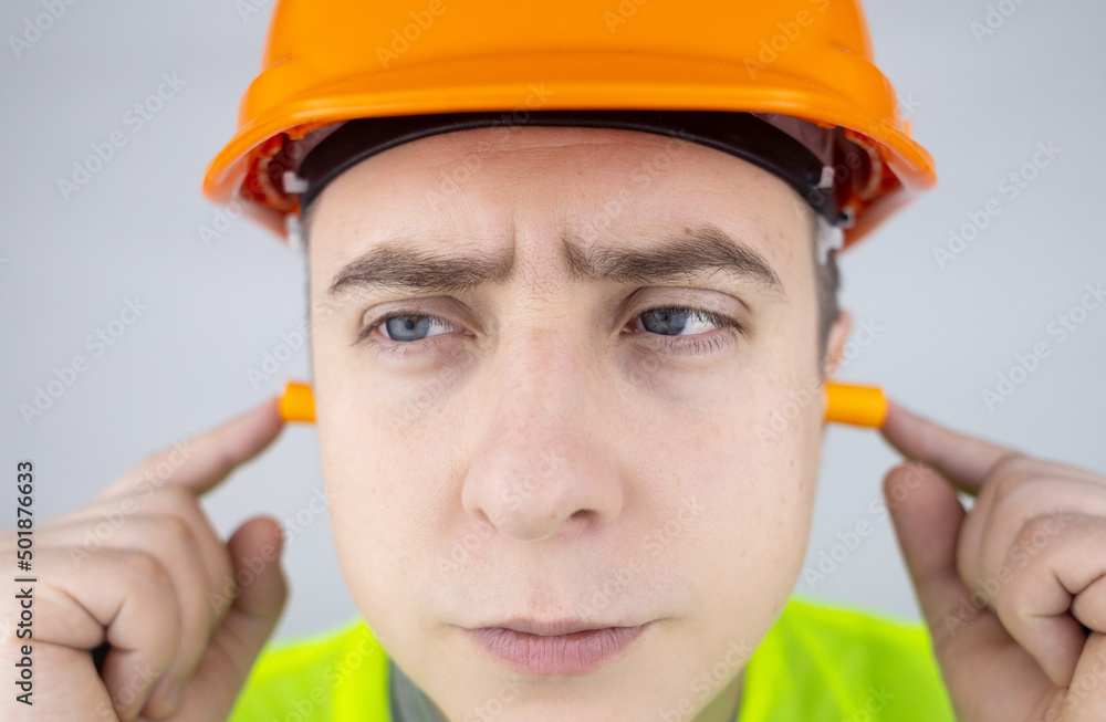 A worker in a hard hat and green vest puts on ear plugs. Industrial ...