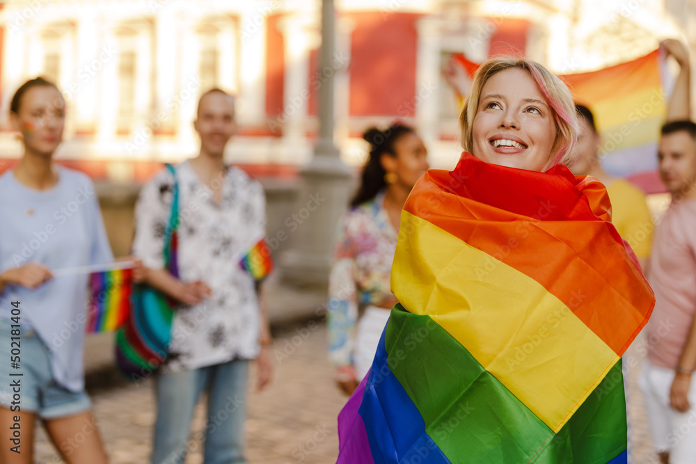 Young lesbian woman wrapped in rainbow flag smiling during pride parade ...