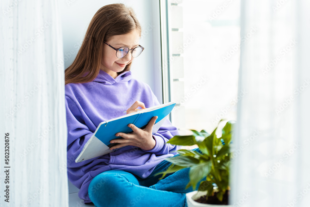 Portrait of smiling schoolgirl sitting near the window, writing essay ...