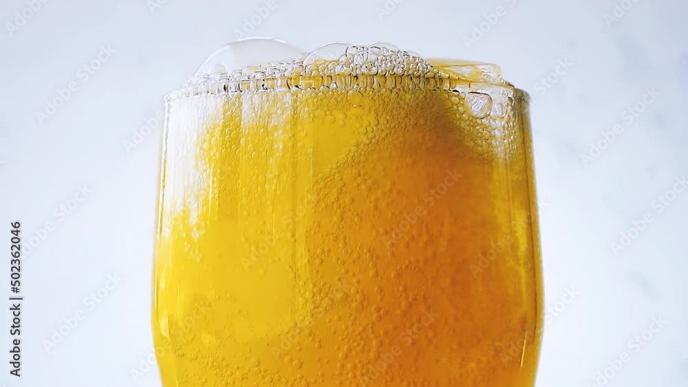 Orange soda is pouring into a glass with ice cube close-up on a white ...