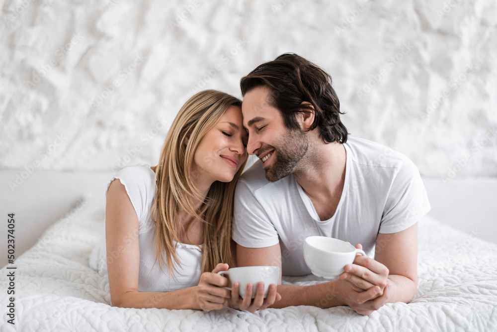Romantic young couple lying on bed with cups of coffee, cuddling ...