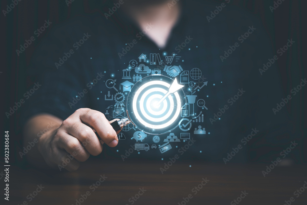 Businessman holding magnifier glass with virtual dartboard arrow and ...