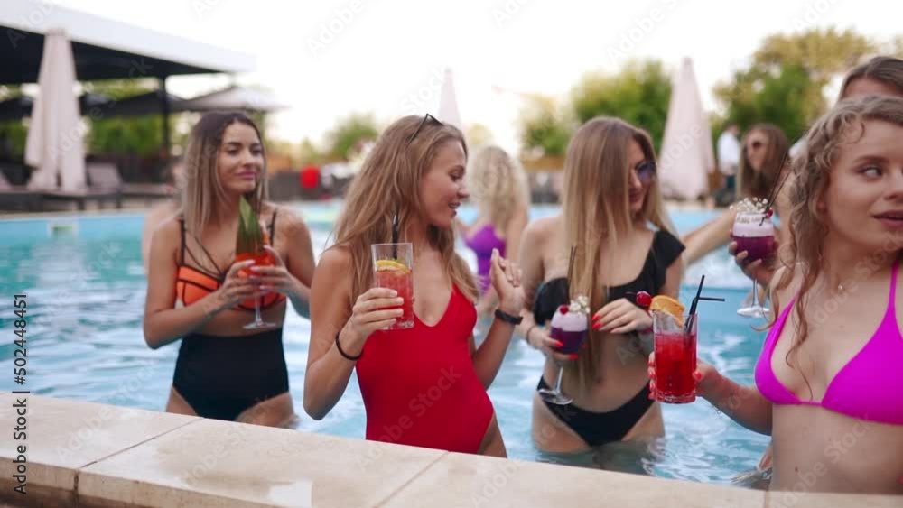 Girls in bikini have pool party with cocktails in swimming pool. Women relaxing clinking glasses ...