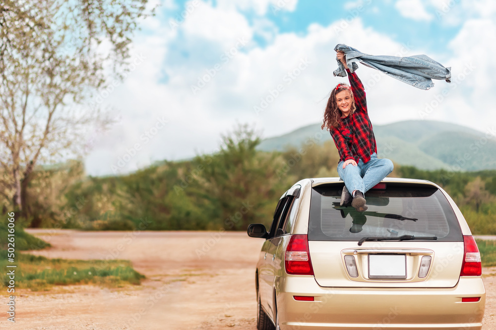 Happy teen girl is sitting on the roof of a car, joyfully raising her ...
