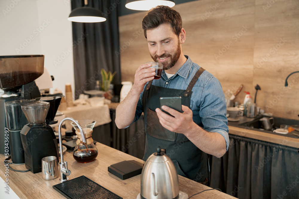 Pleasant barista drinking coffee and scrolling through social media ...
