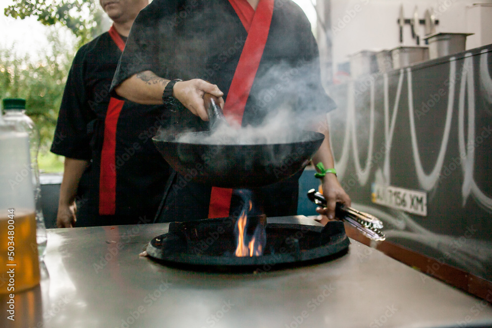 cook fries vegetables in a wok pan on fire. Chef is stirring vegetables ...