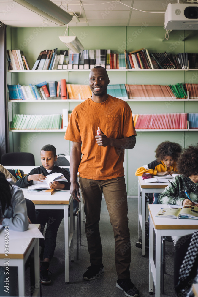 Portrait of confident teacher showing thumbs up while standing amidst ...
