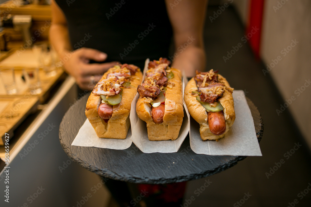 cook in black gloves holds hot dog. The cook prepares a hot dog, adding ...