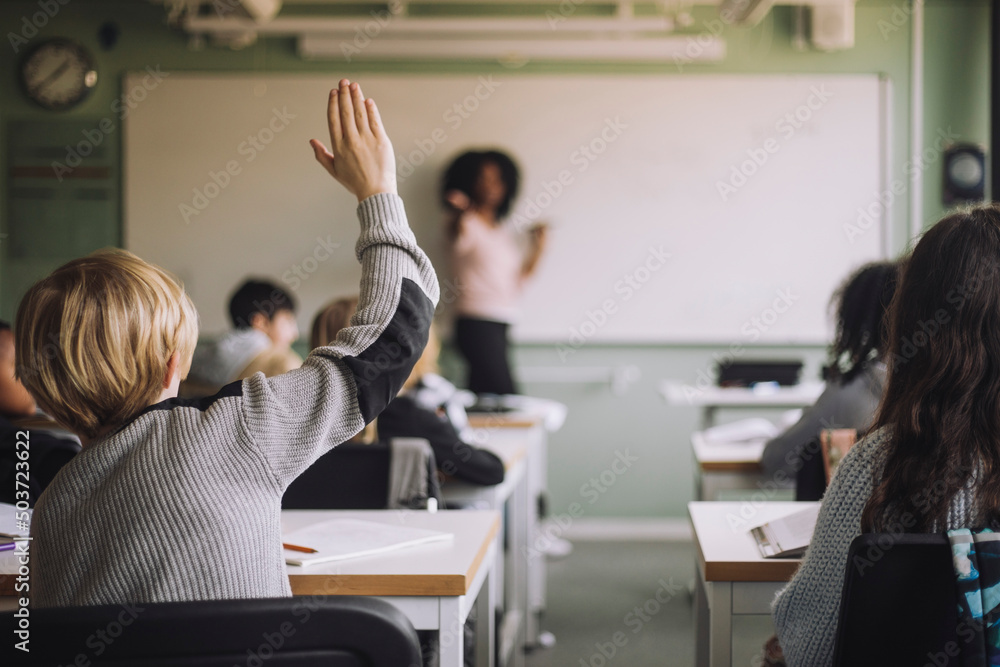 Rear view of student raising hand while teacher teaching in classroom ...