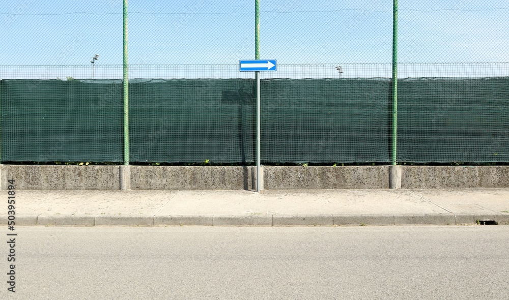 Road directional arrow sign on a concrete sidewalk. Wire mesh fencing ...