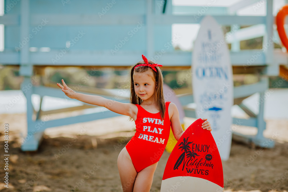 Pretty little girl in red bikini posing with small surfboard like a model on the beach against ...