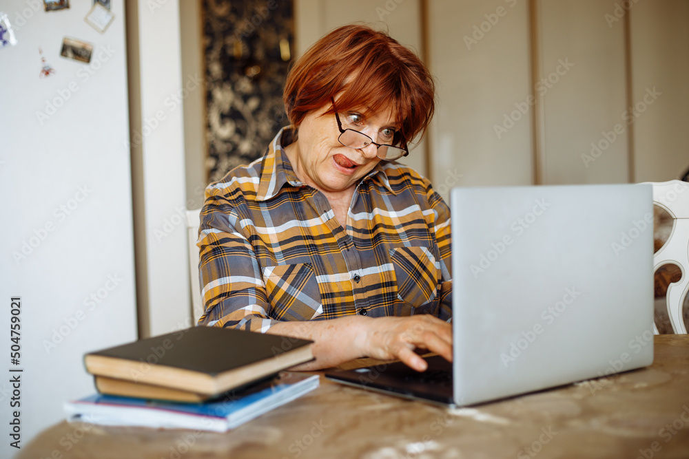 Mature lady in glasses typing on laptop at table in room and practicing ...