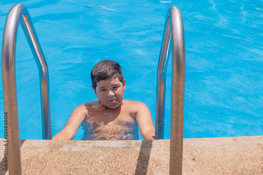 boy in the pool on summer vacation with a funny face at the edge of the pool holding on to the ...