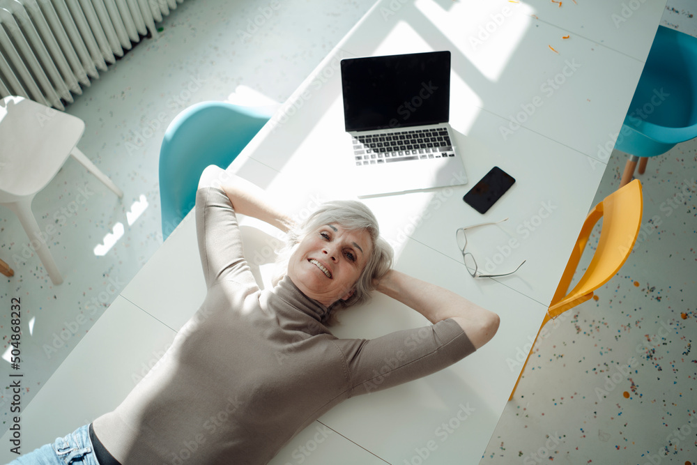 Smiling senior businesswoman with hands behind head lying on desk in ...
