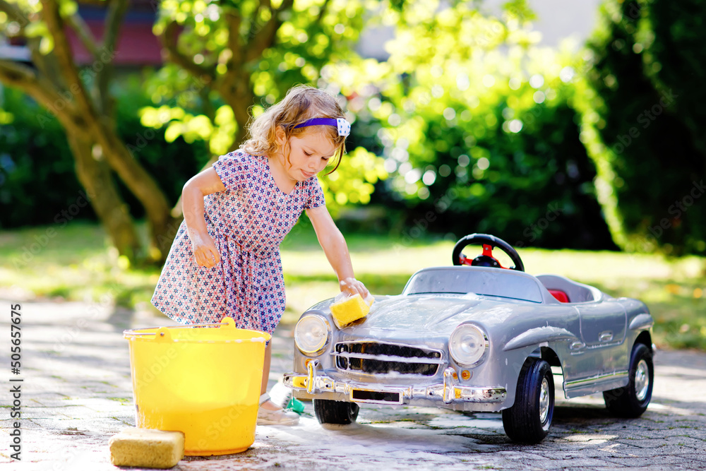 Cute gorgeous toddler girl washing big old toy car in summer garden ...