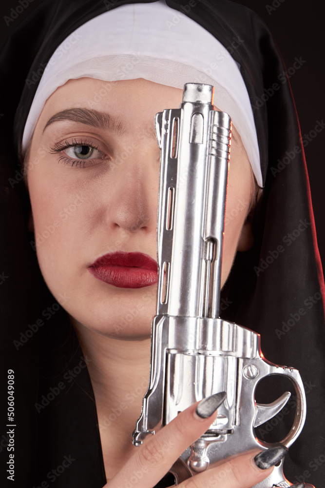 Close-up portrait of a nun holds silver revolver gun up. Young female ...