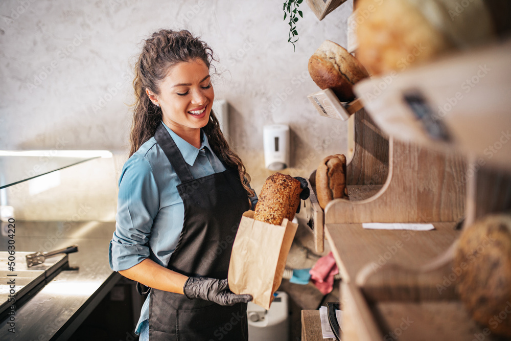 Female worker working in bakery. She packs the bread into a paper bag ...