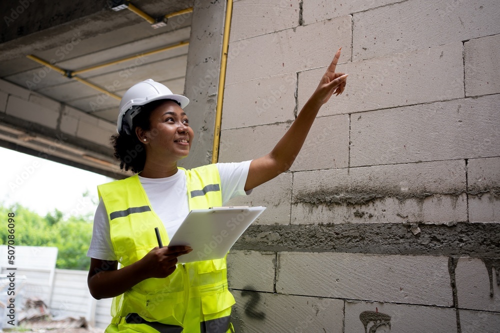 African American woman worker at construction site Wearing a hard hat,vest, holding clipboard ...