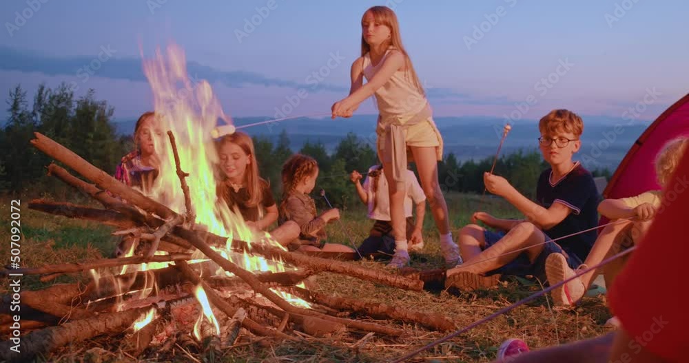 Side view of group of kids sitting around big campfire with roasted meal at twilight. Happy ...