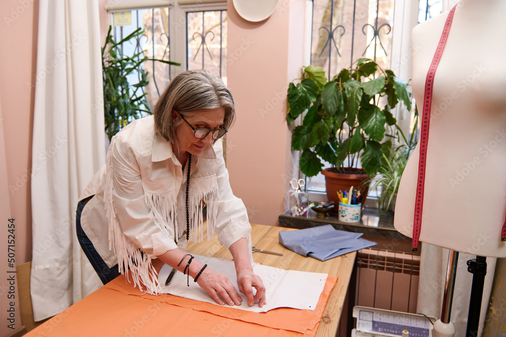 Mature female tailor works with pattern and fabric, creating new ...
