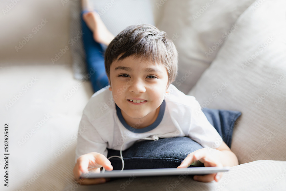 High key portrait young boy playing game on tablet sitting on sofa with ...