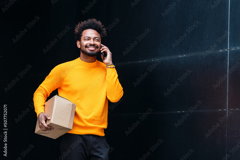 Smiling delivery man holding box talking on mobile phone in front of ...
